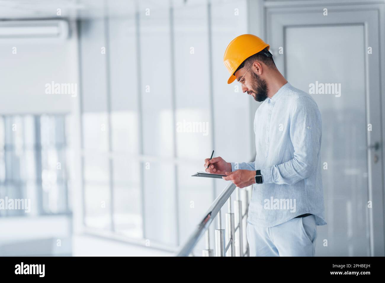 Holding notepad. Engineer in white clothes and orange protective hard hat standing and working ...