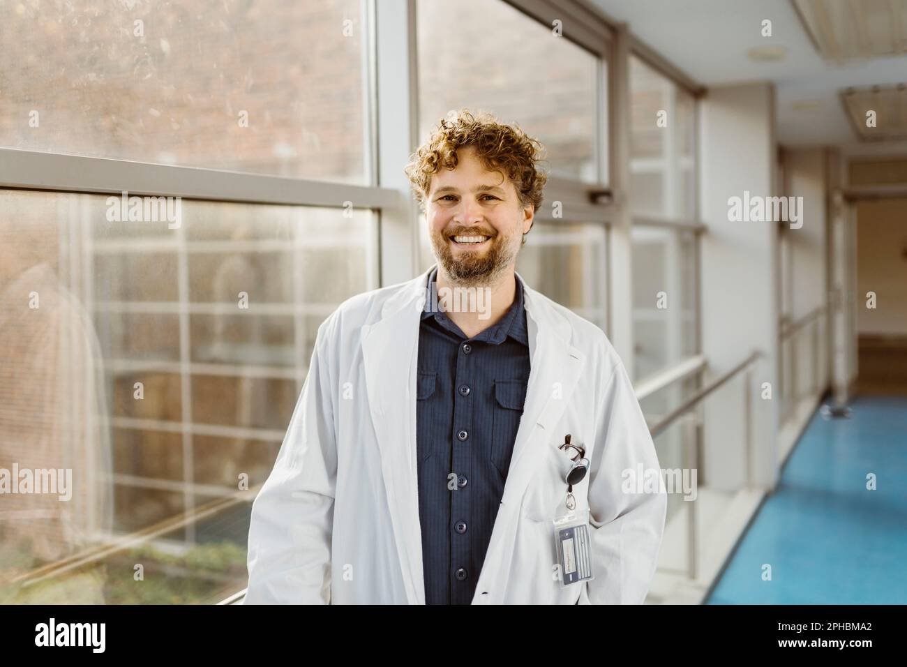 Portrait of smiling mature doctor wearing lab coat standing by window ...