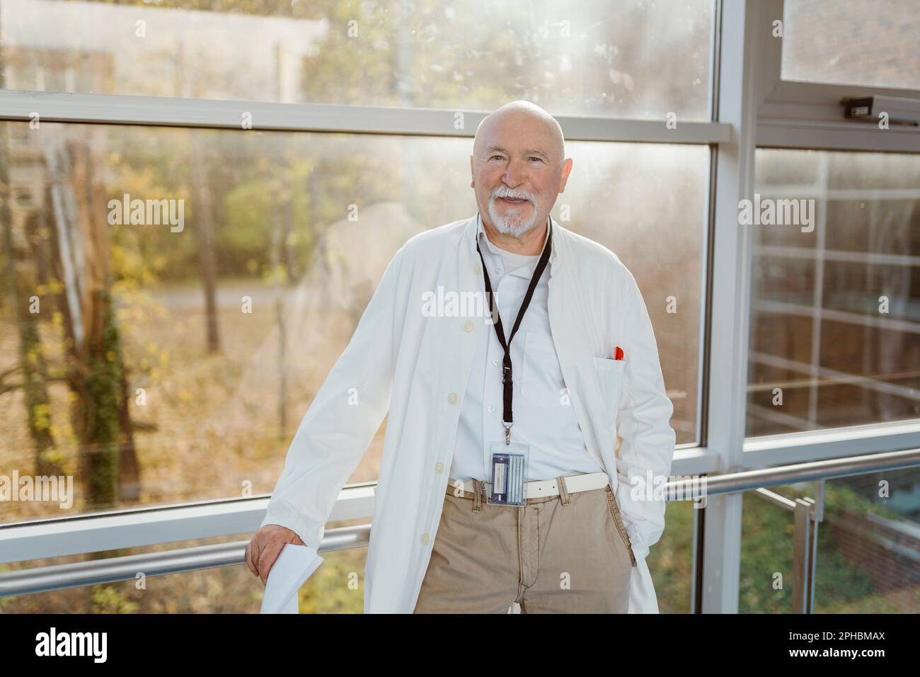 Portrait of senior male doctor leaning on railing against window at ...