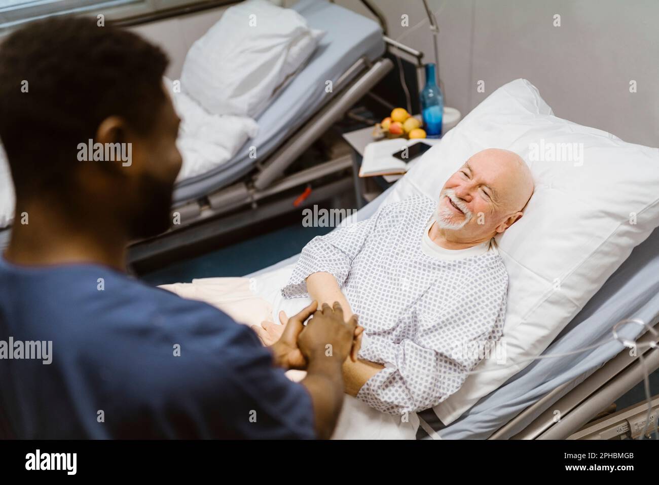 Male Nurse Holding Hands Of Smiling Senior Patient Lying On Bed In