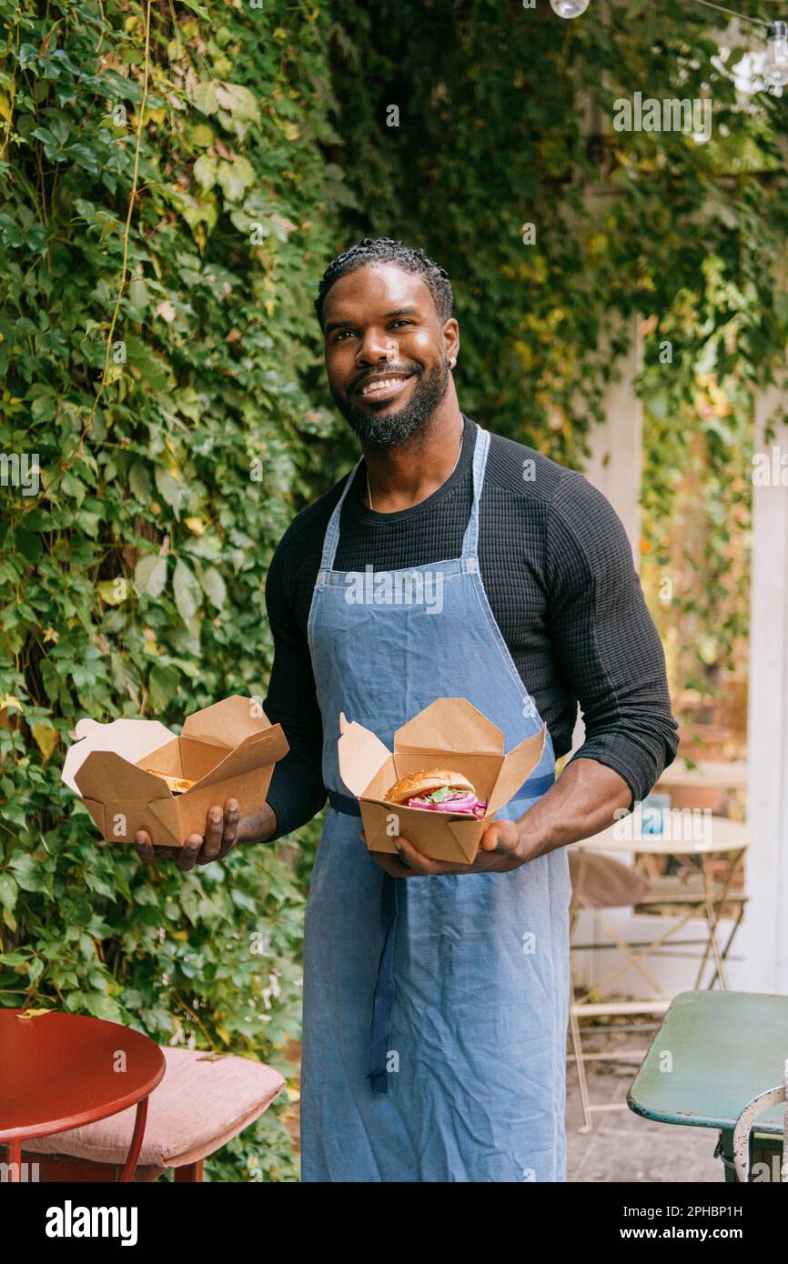 Portrait of smiling male cafe owner holding food in take away boxes ...