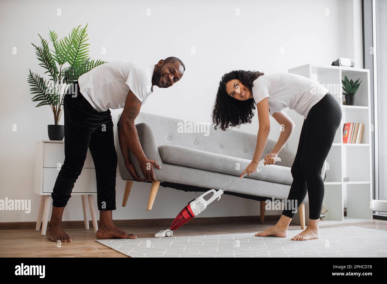 Happy interracial woman using modern cordless vacuum for cleaning underneath of couch that ...