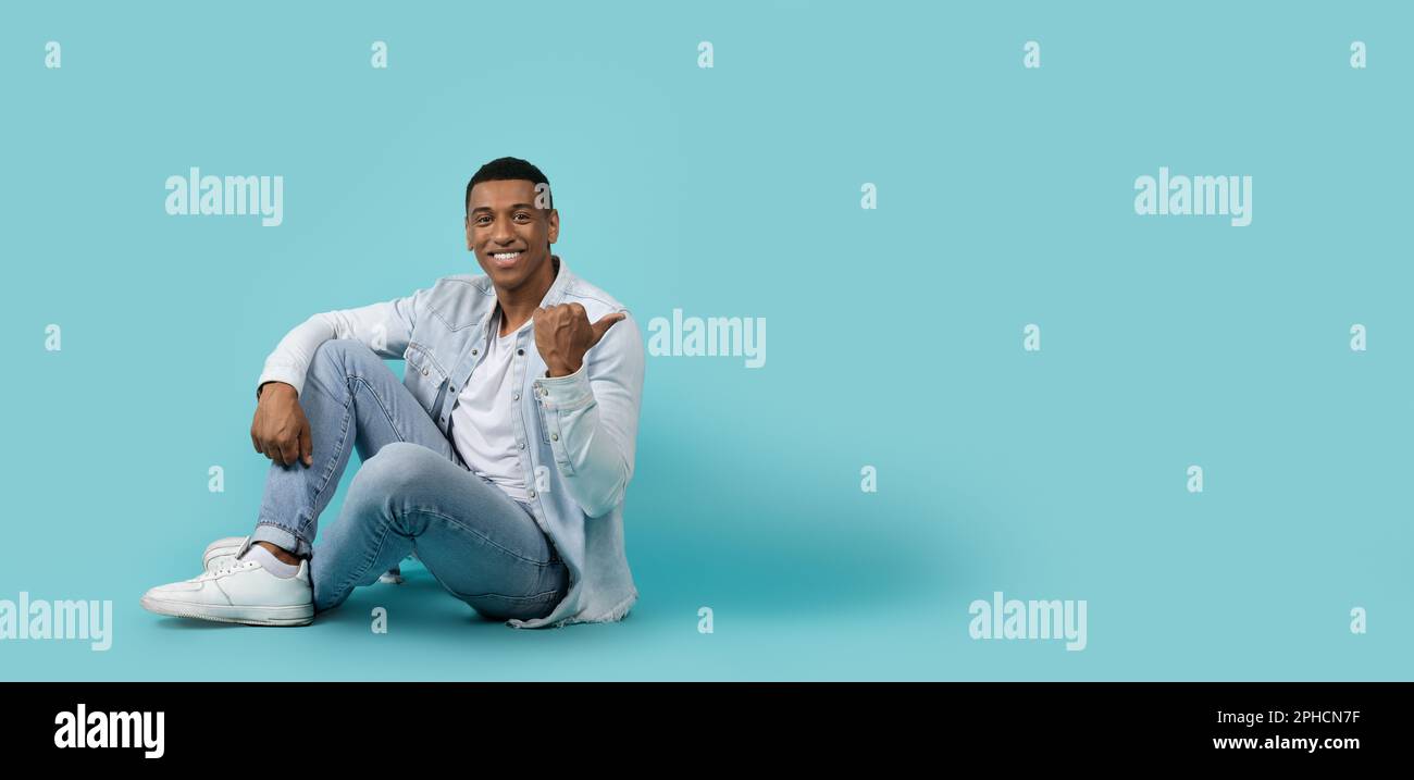 Cheerful millennial black guy sitting on floor, pointing finger at ...