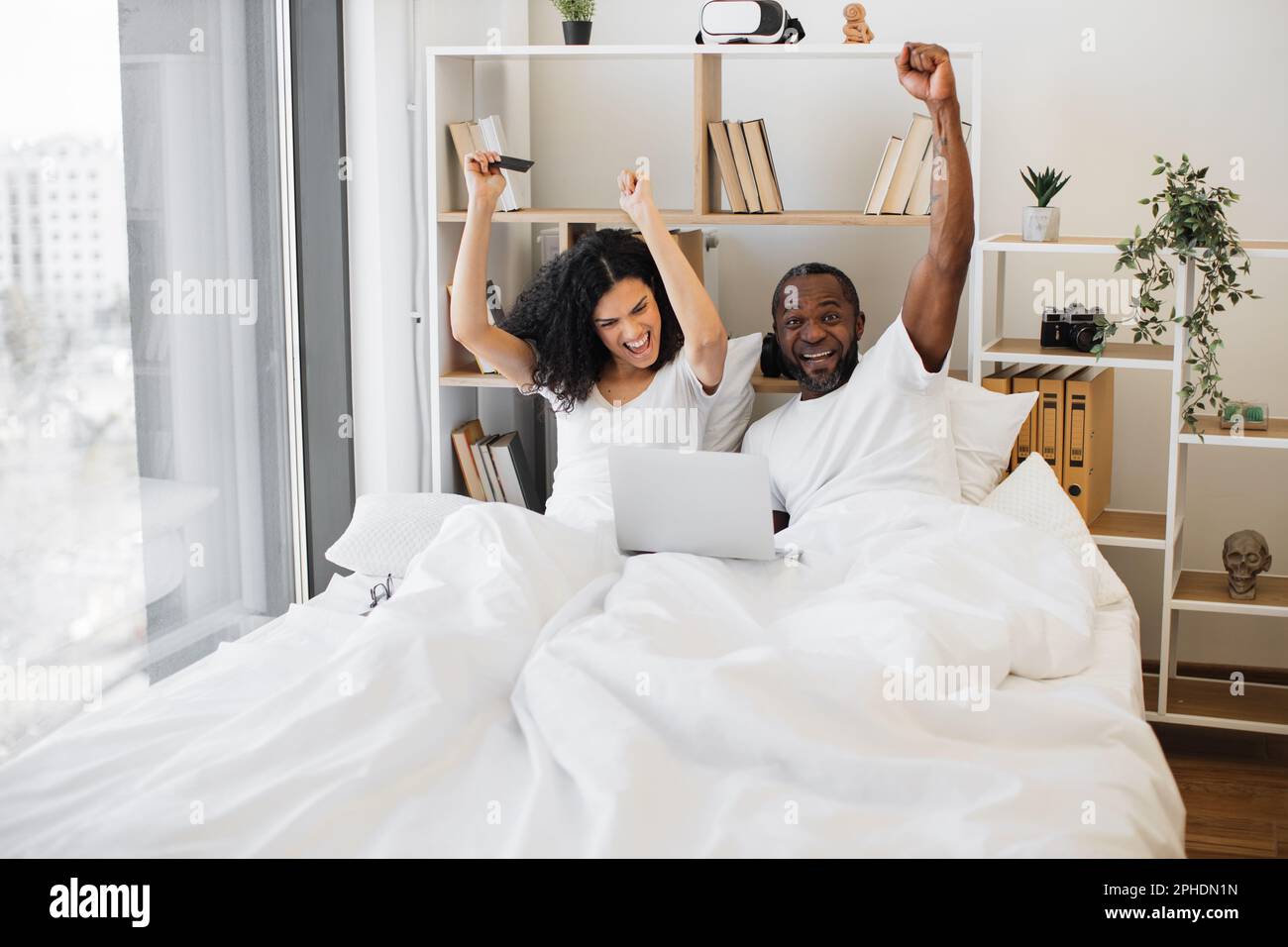 Emotional african american family of two raising hands in excitement while sitting in bed with ...