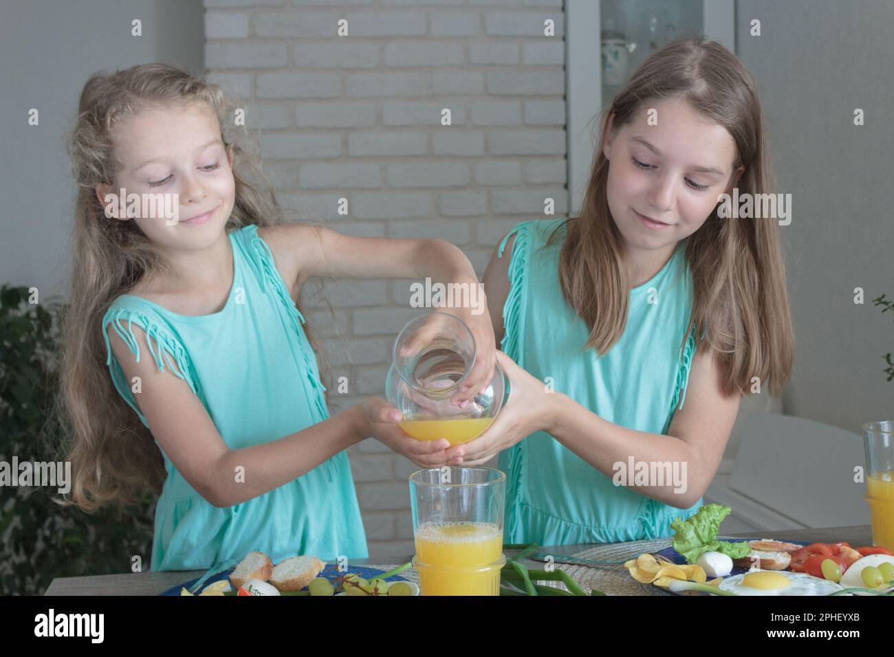 Two girls with long hair in identical dresses are pouring orange juice ...