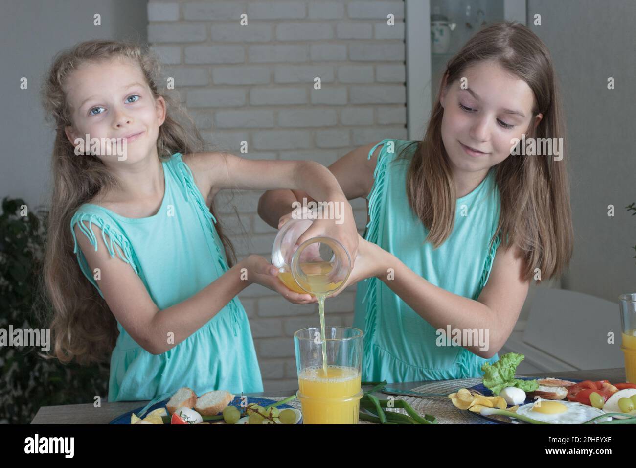 Two girls with long hair in identical dresses are pouring orange juice ...