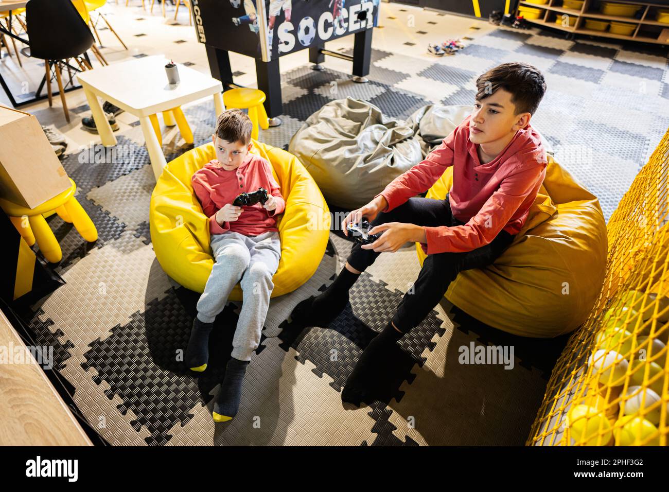 Two brothers playing video game console, sitting on yellow pouf in kids ...