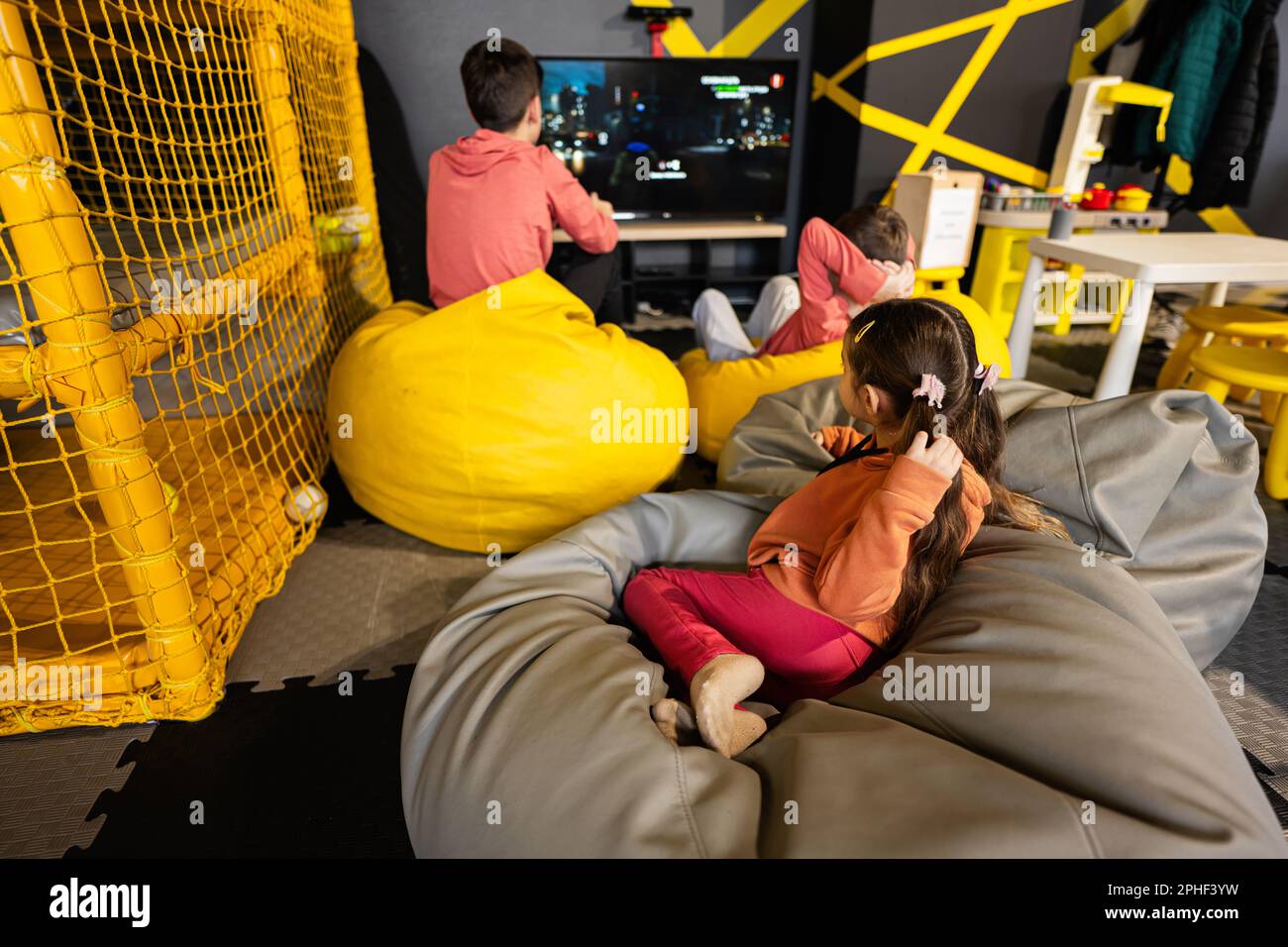 Three children playing video game console, sitting on yellow pouf in ...