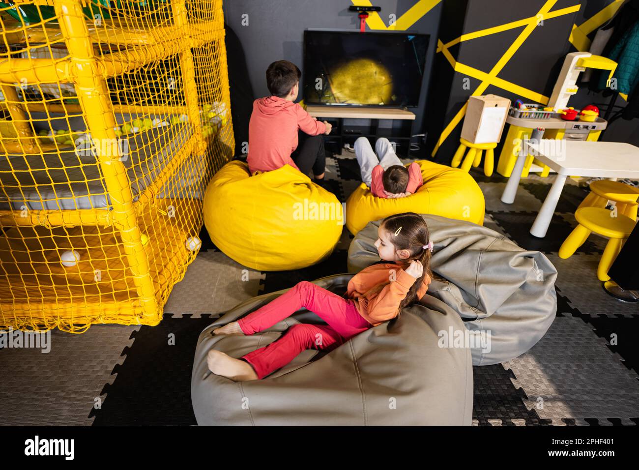 Three children playing video game console, sitting on yellow pouf in ...