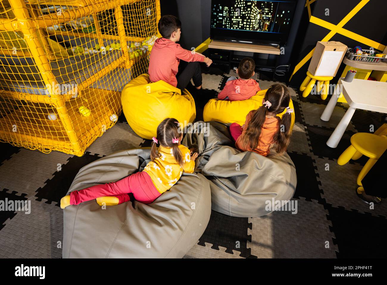 Four children playing video game console, sitting on yellow pouf in ...