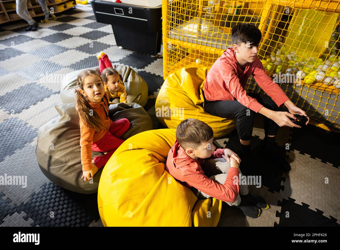Four children playing video game console, sitting on yellow pouf in ...