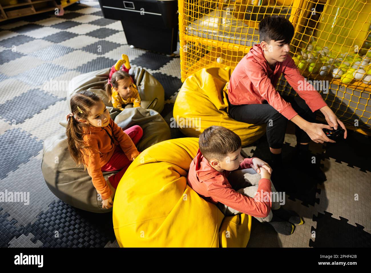 Four children playing video game console, sitting on yellow pouf in ...