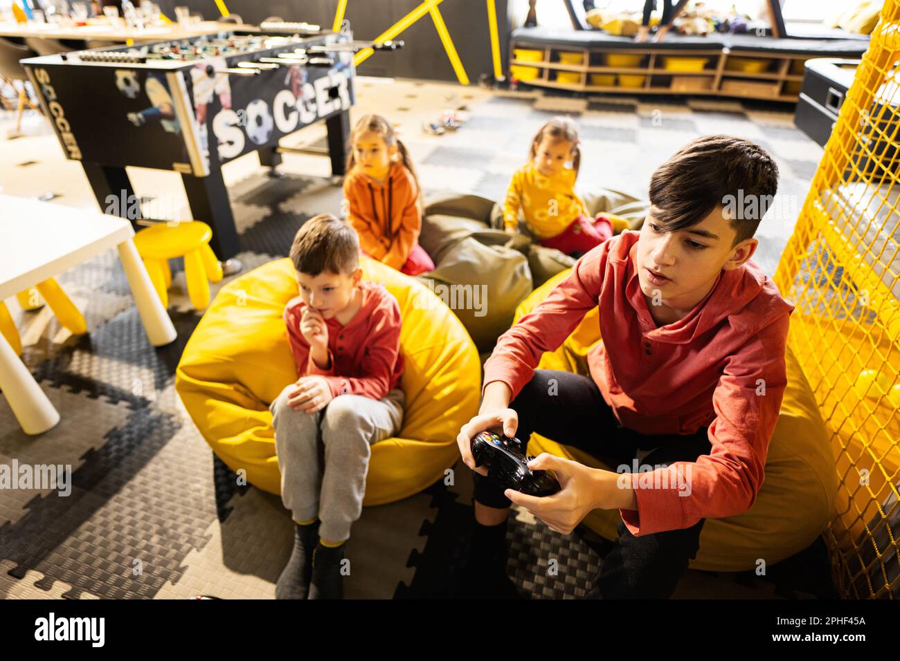 Four children playing video game console, sitting on yellow pouf in ...