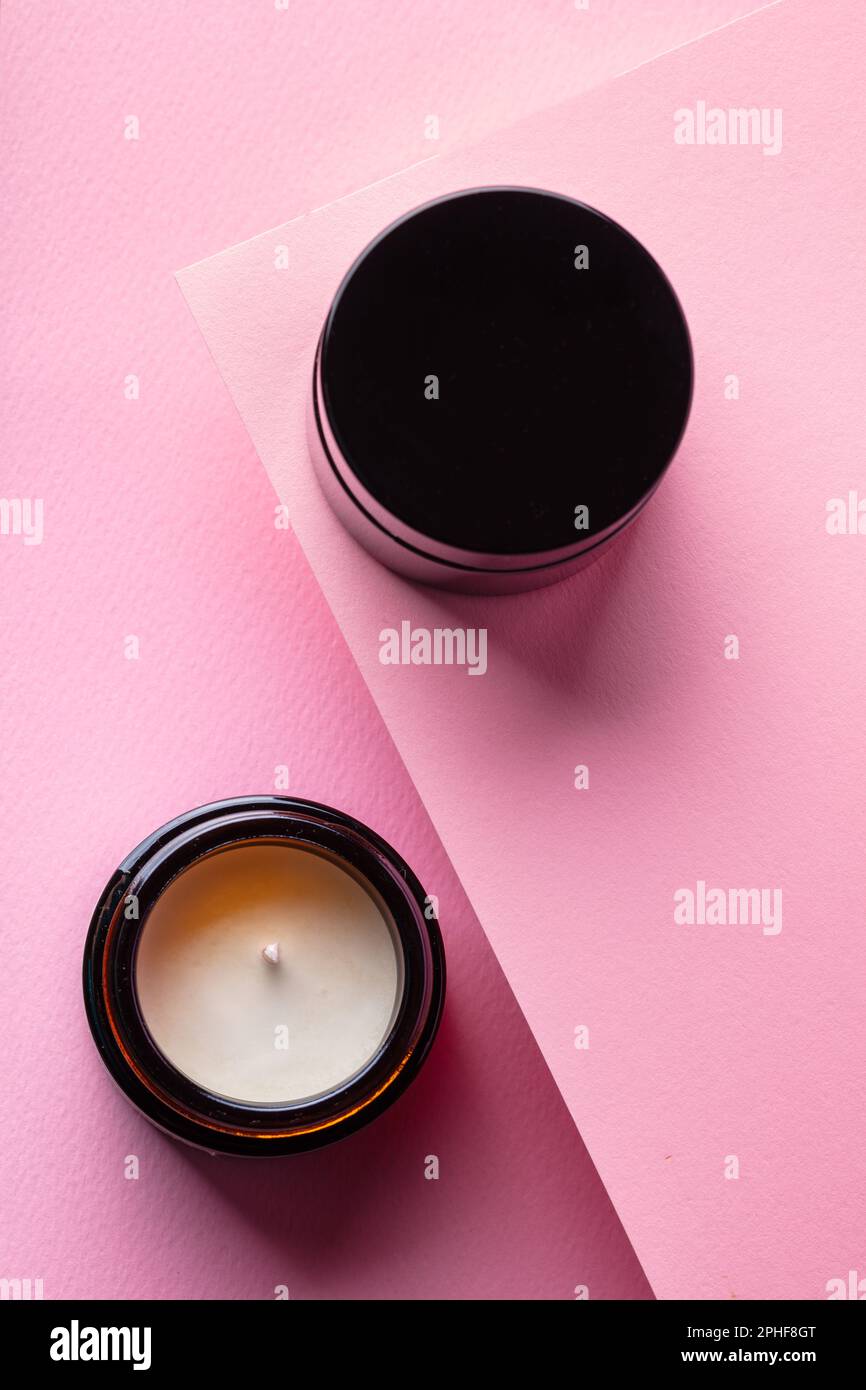 A black container and a lit candle are displayed on a pink background ...