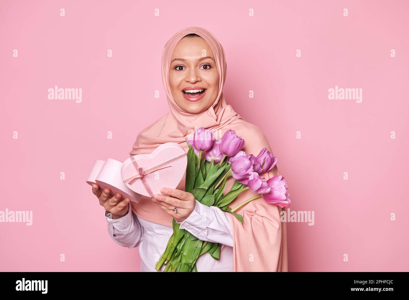 Cheerful Arab lady in pink hijab, holding a heart shaped gift box and ...