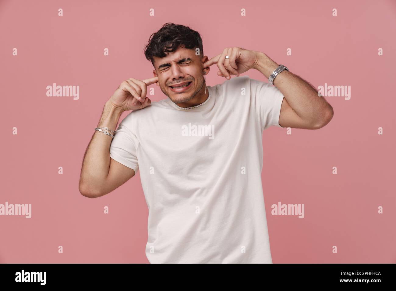 Brunette young man grimacing while plugging his ears isolated over pink ...