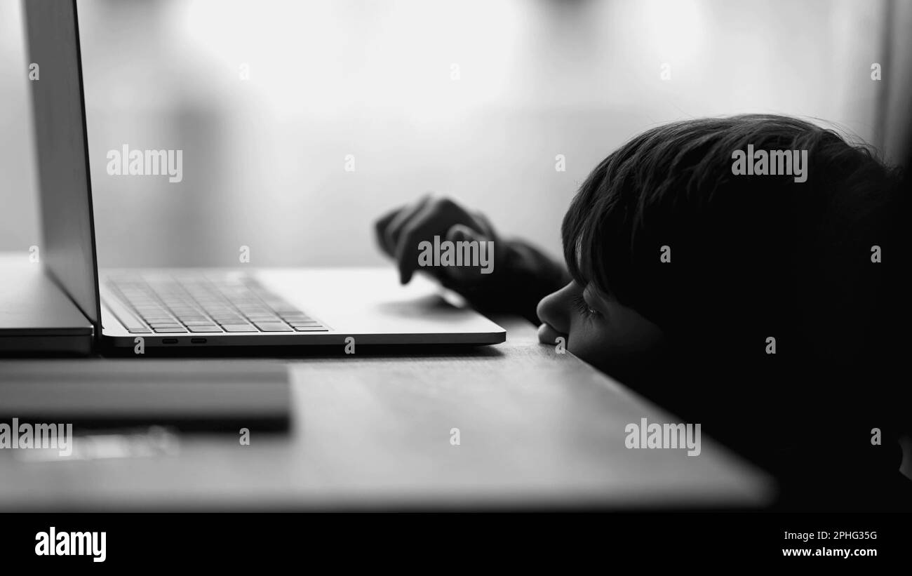 One bored little boy leaning on table and using computer laptop feeling ...