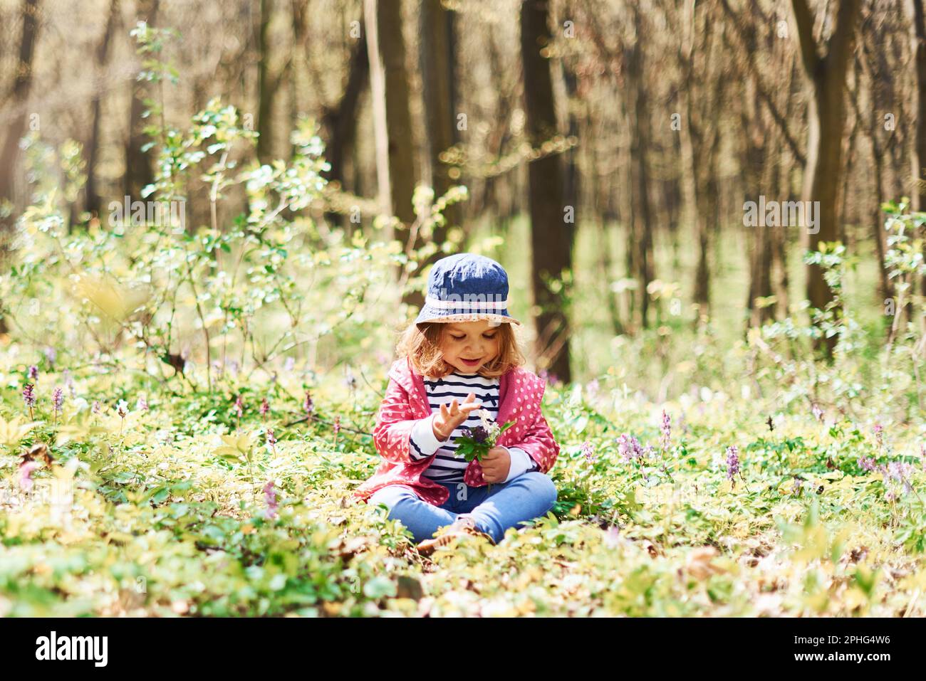 Happy little girl in blue hat have walk in spring forest at daytime ...