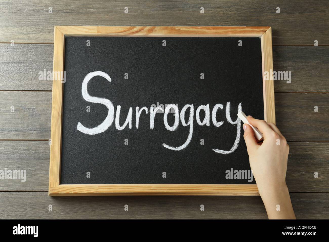 Woman writing word Surrogacy on small blackboard at wooden table, top ...