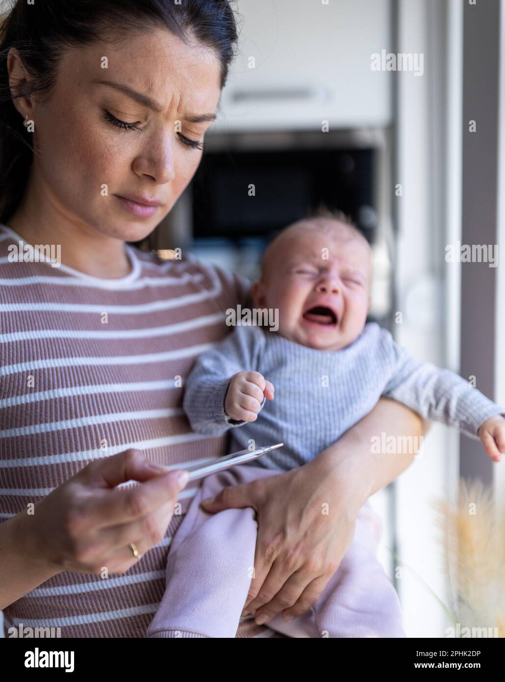 Worried mother holding crying baby in one hand at home and looking at thermometer for fever ...