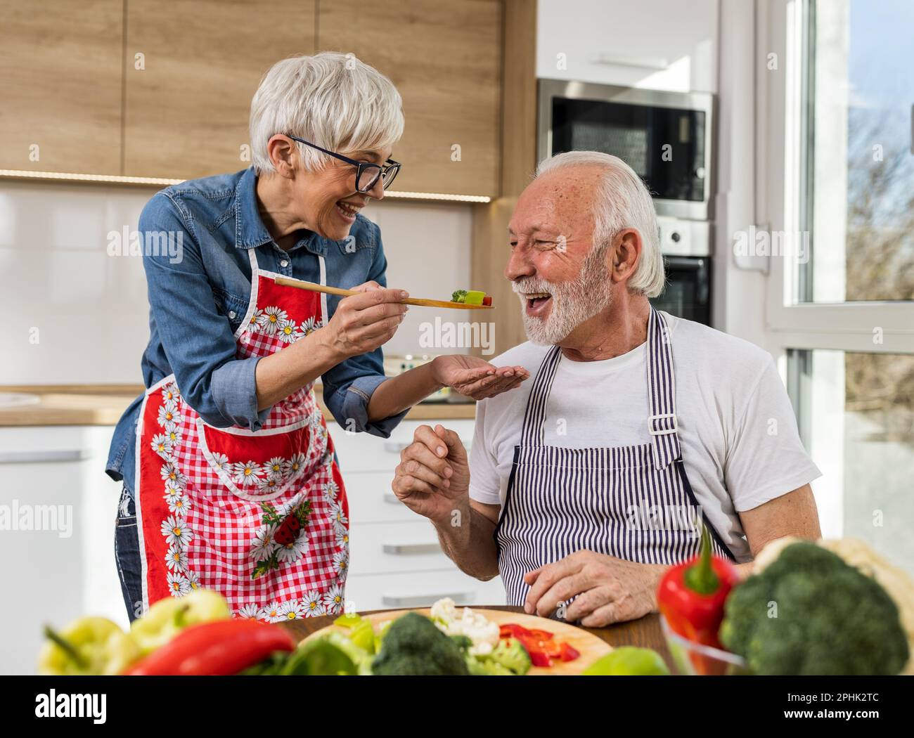 Cheerful senior couple preparing food, man tasting cooked vegetable ...