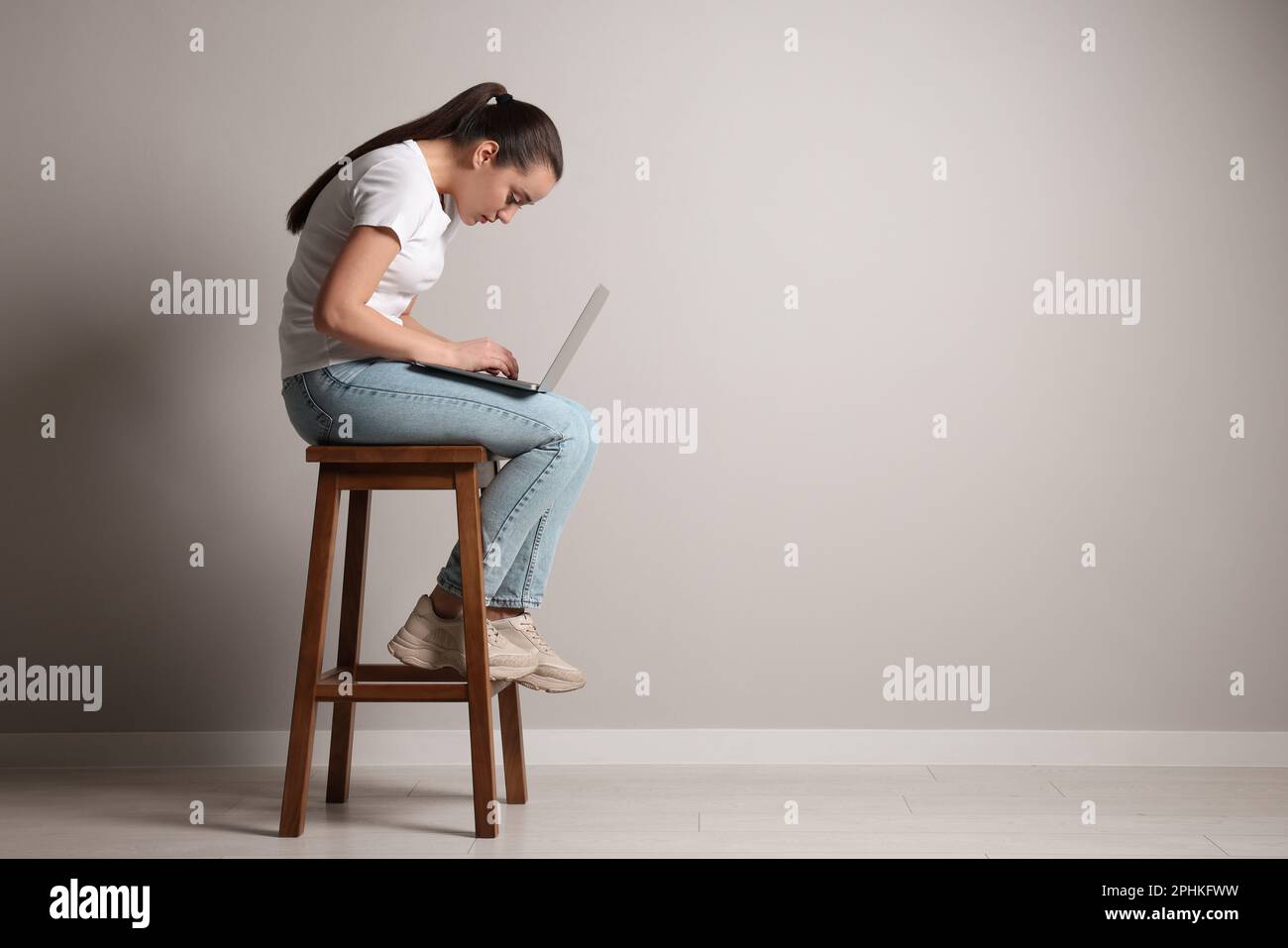 Young woman with poor posture using laptop while sitting on stool near ...