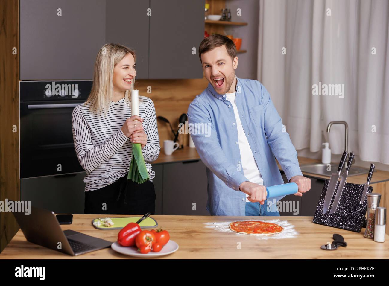 Happy young husband and wife laugh and sing using kitchen appliances ...