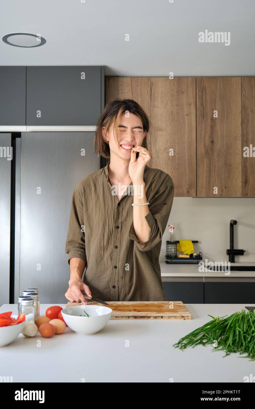 Smiling asian young man biting chinese chive at kitchen while cooking ...