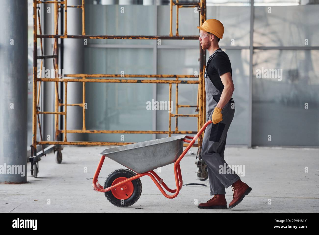 Walks with wheelbarrows. Man in grey uniform works indoors in modern big office at daytime Stock ...