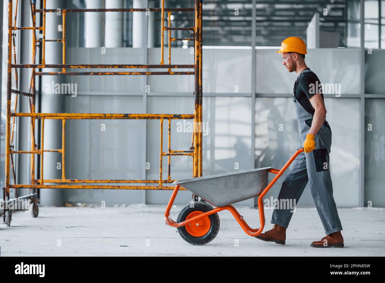 Walks with wheelbarrows. Man in grey uniform works indoors in modern big office at daytime Stock ...