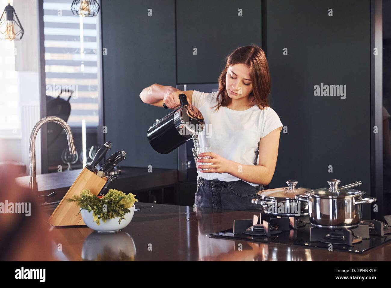 Girl in white shirt standing indoors in kitchen and poring water into ...