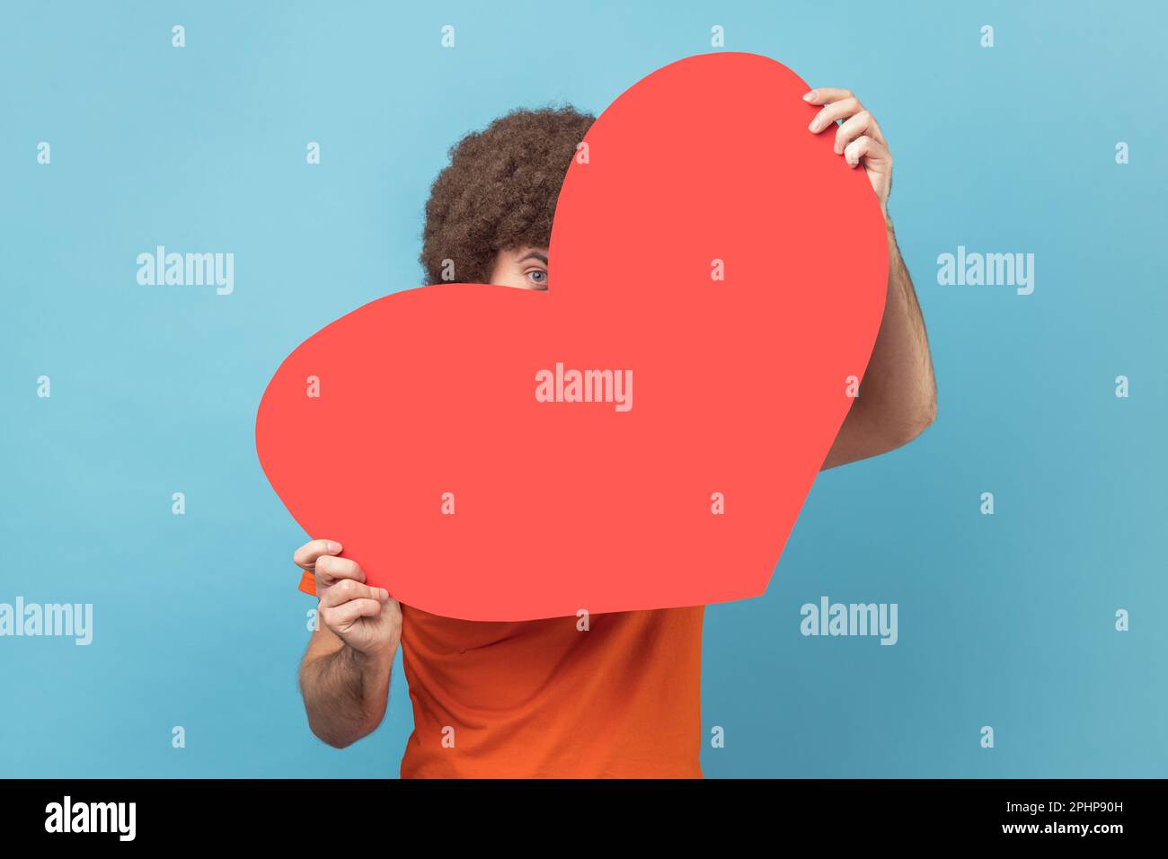 Portrait of man hiding face behind big red heart and looking at camera ...