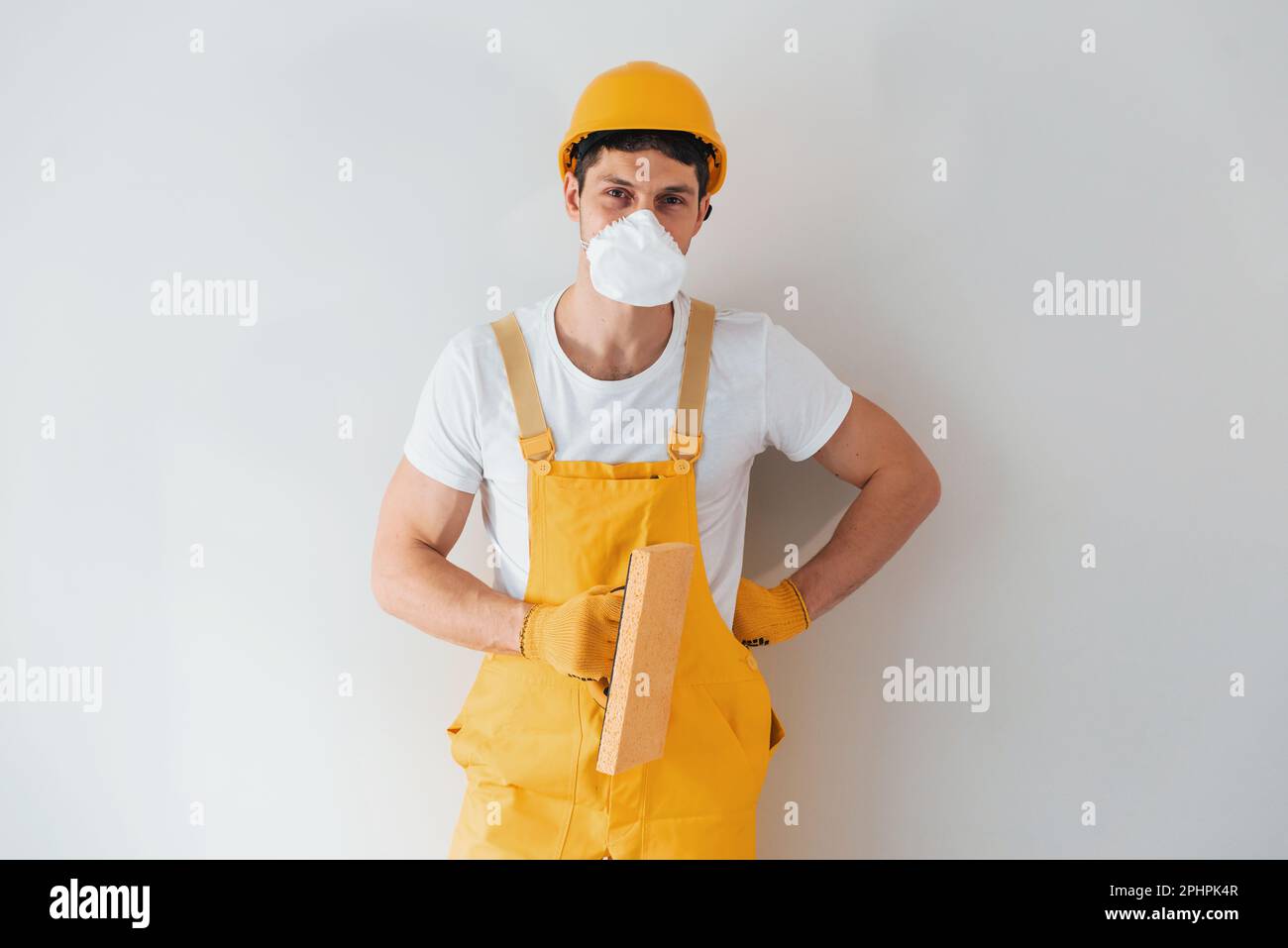 Handyman in yellow uniform and protective mask standing against white ...