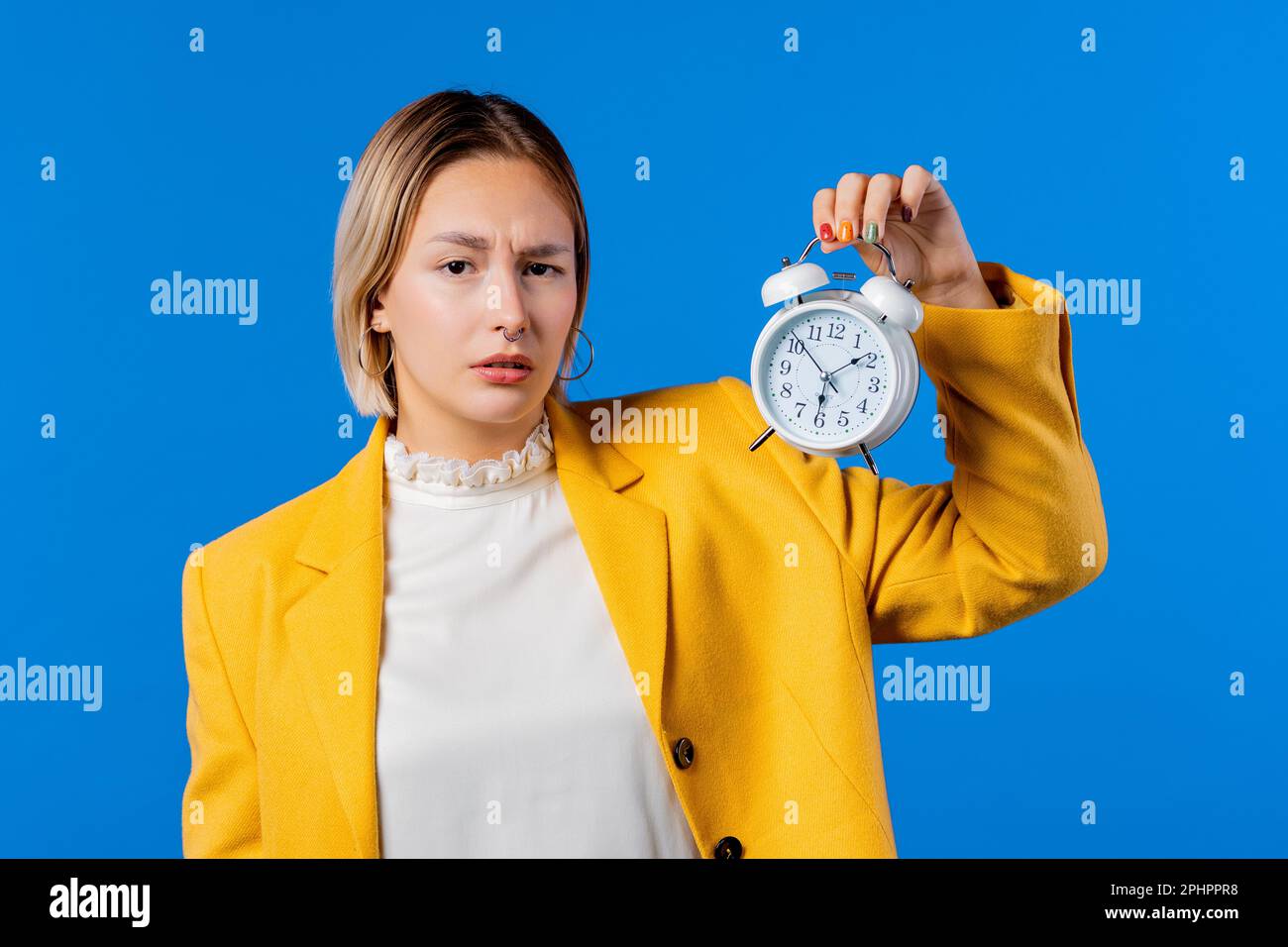 Confused woman holding alarm clock in hand. Blue background. Early 6 o ...