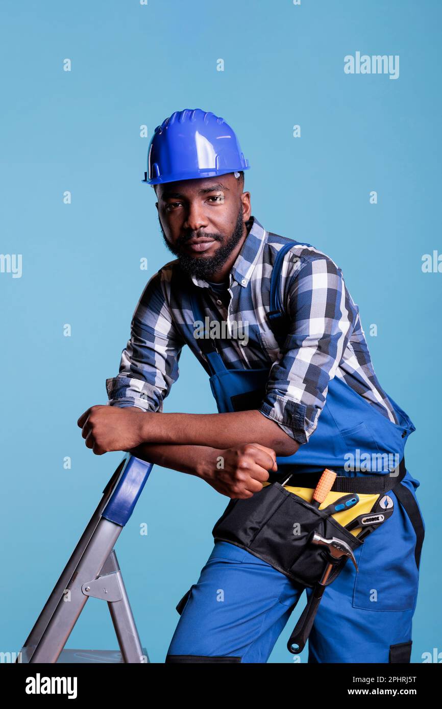 Builder looking calm leaning against ladder in studio shot against blue ...