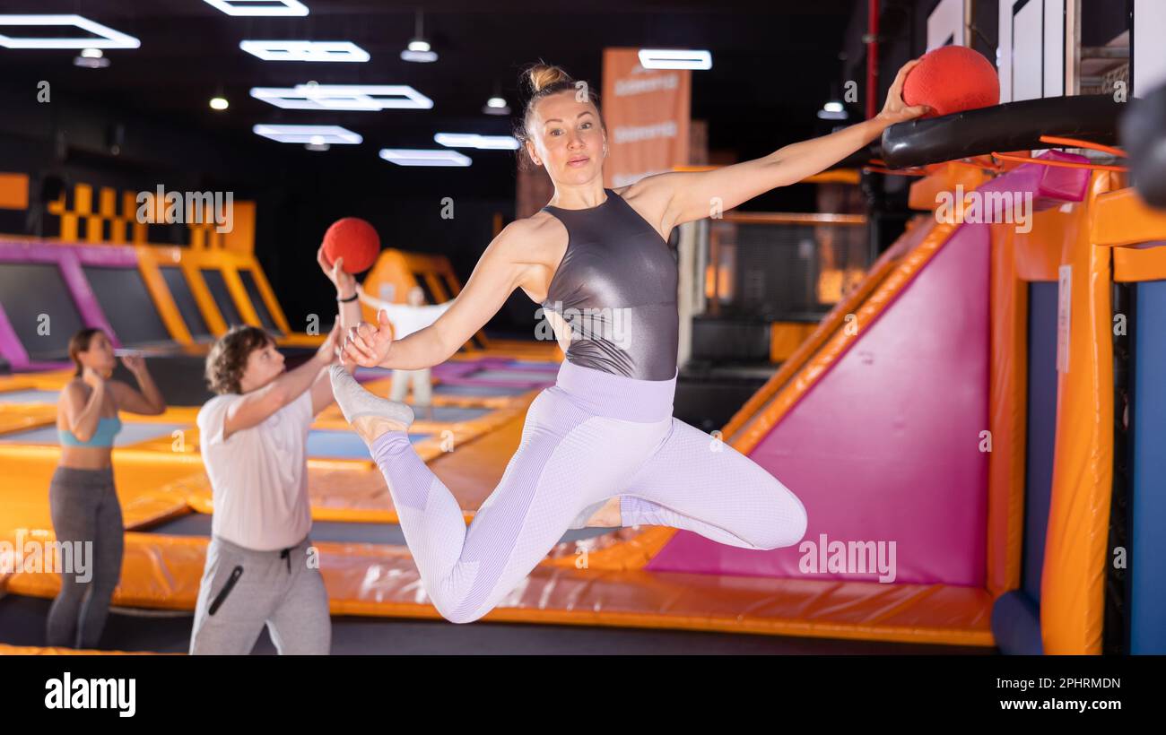 Middle-aged woman throwing ball in basket high jumping in trampoline ...