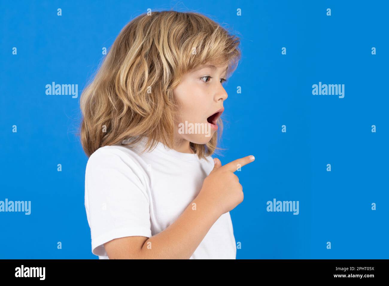 Surprised child looking away, pointing finger on isolated blue ...