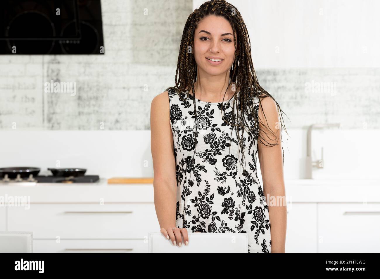In a white kitchen, a young woman with box-braids sighs, smiling ...