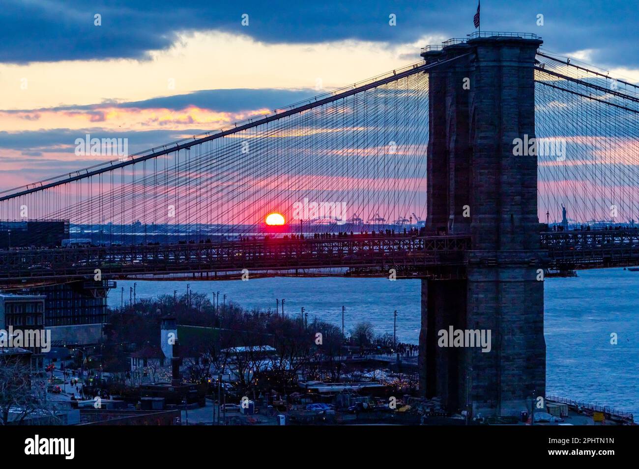 Sunset on the horizon behind the Brooklyn Bridge with crowds of people ...