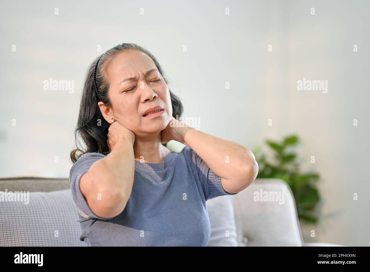 Unhappy elderly Asian woman sits on sofa in her living room suffering ...
