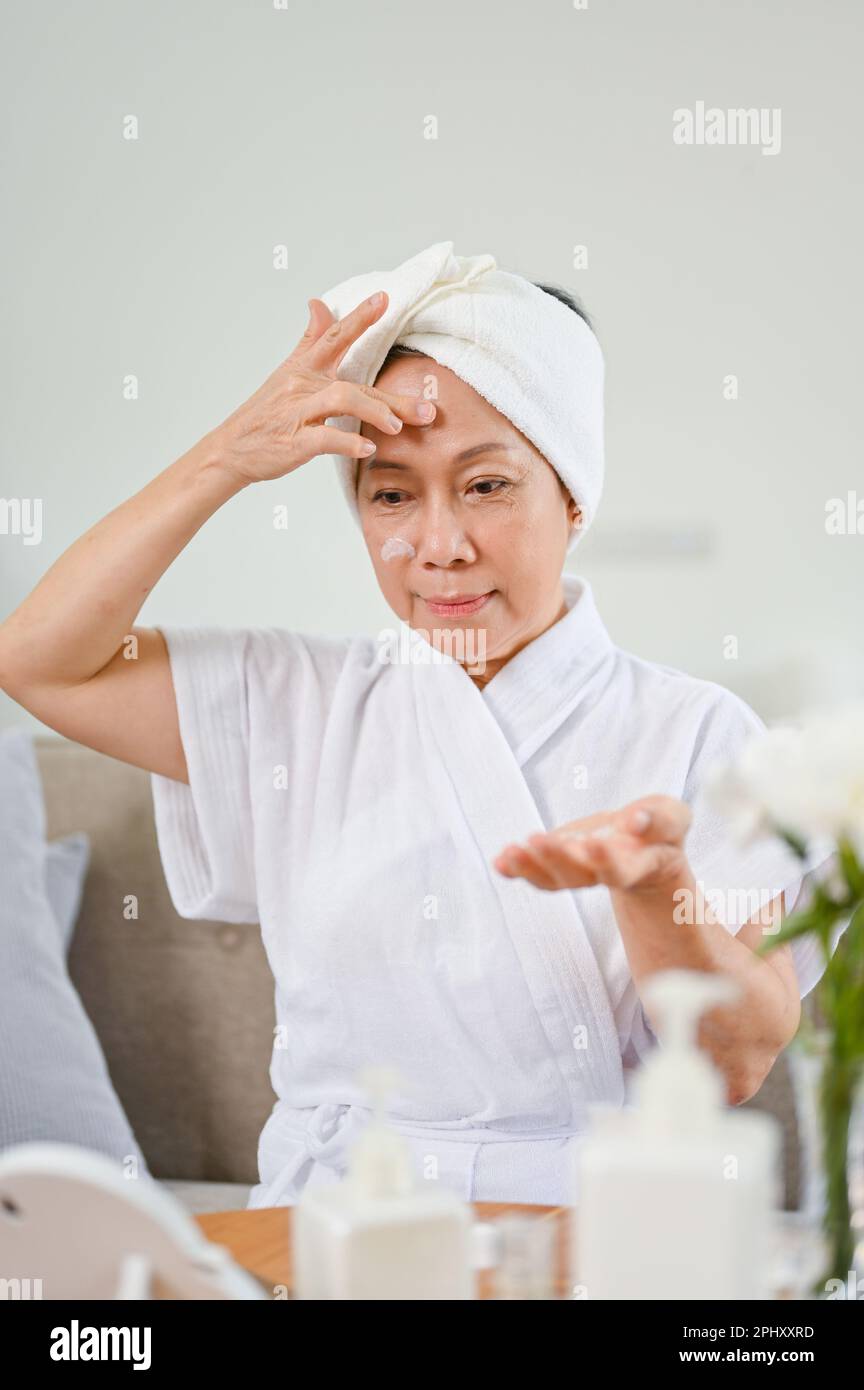 Portrait of attractive Asian-aged woman in bathrobe applying face serum ...