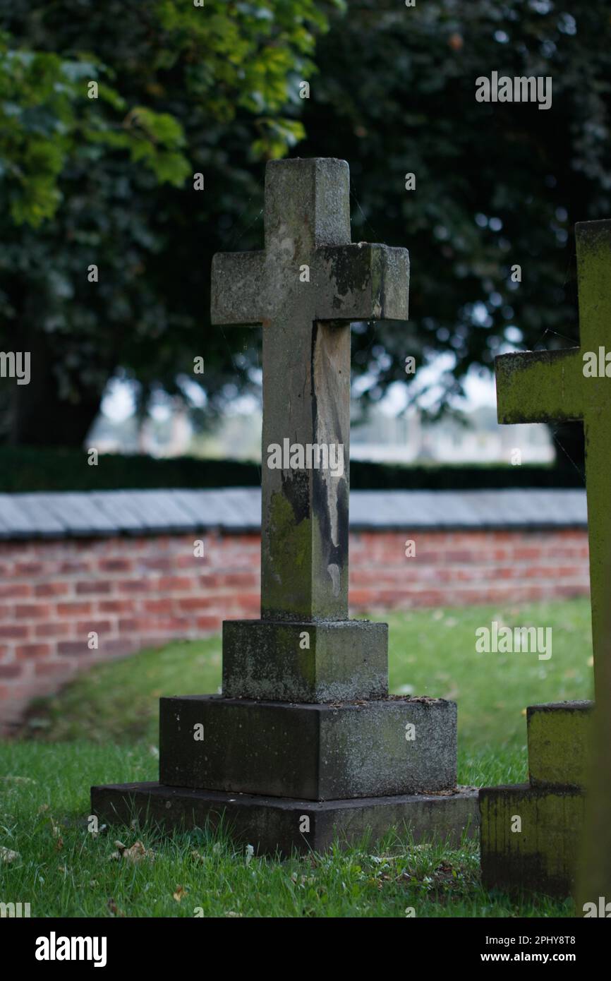 An aged stone cross in a graveyard setting, with a red brick wall in ...