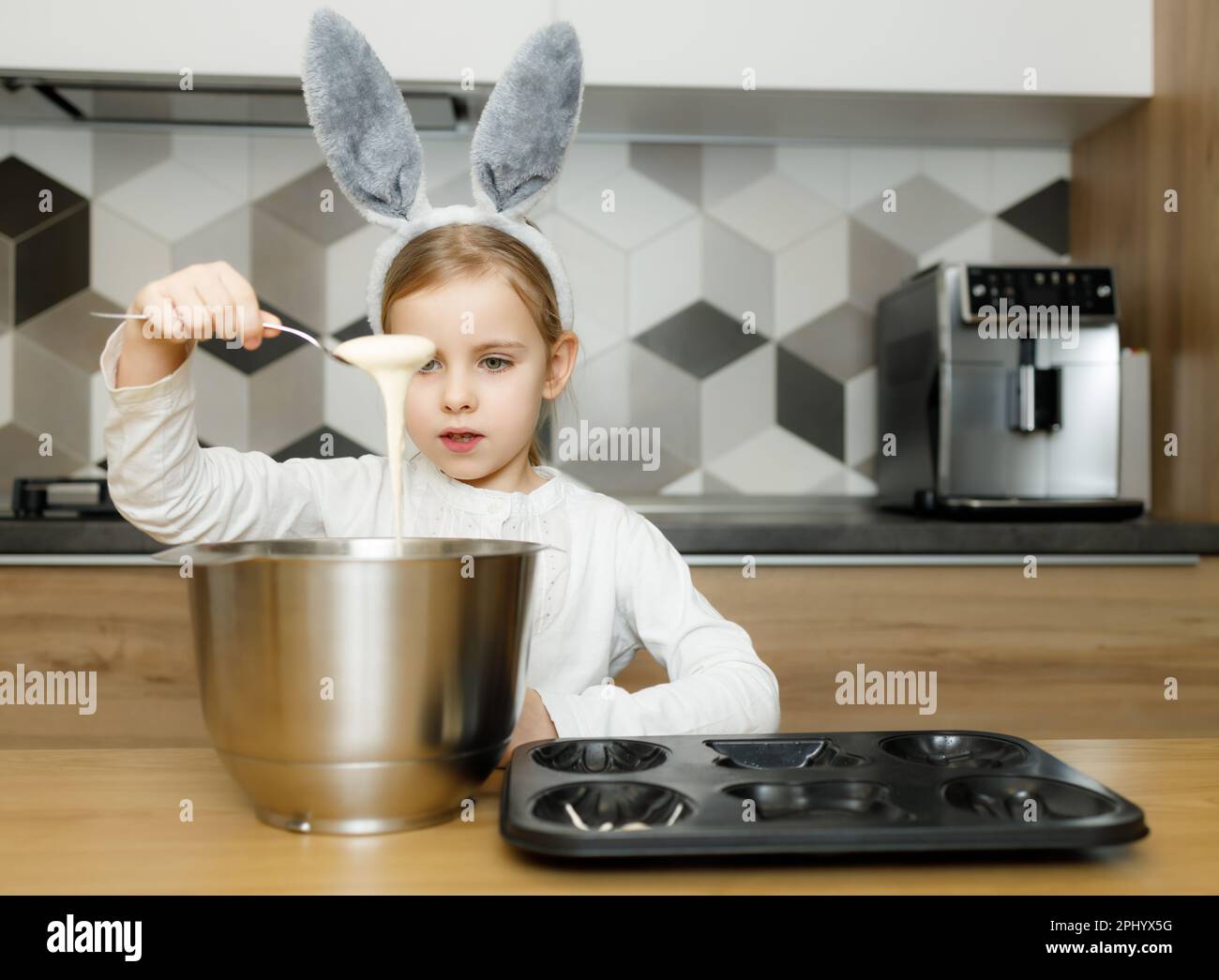 Serious child in bunny ears, little girl carefully pouring raw dough ...