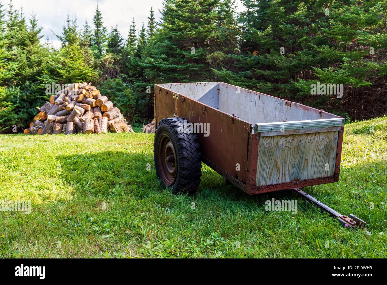 An old utility trailer in a field with a pile of wood in the background ...
