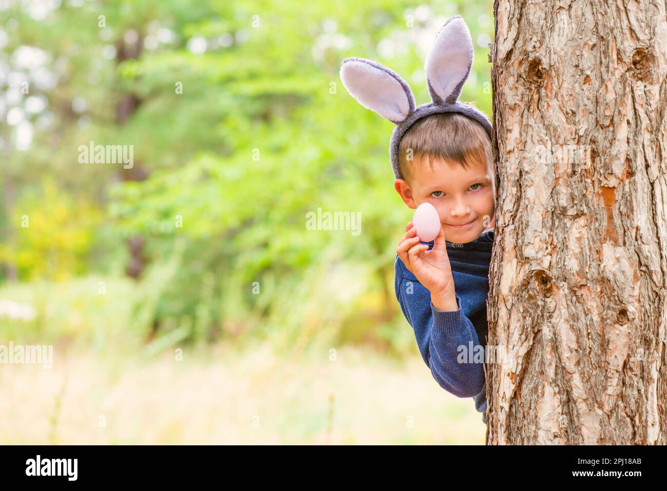 Cute boy with bunny ears standing behind tree and holding easter egg in ...