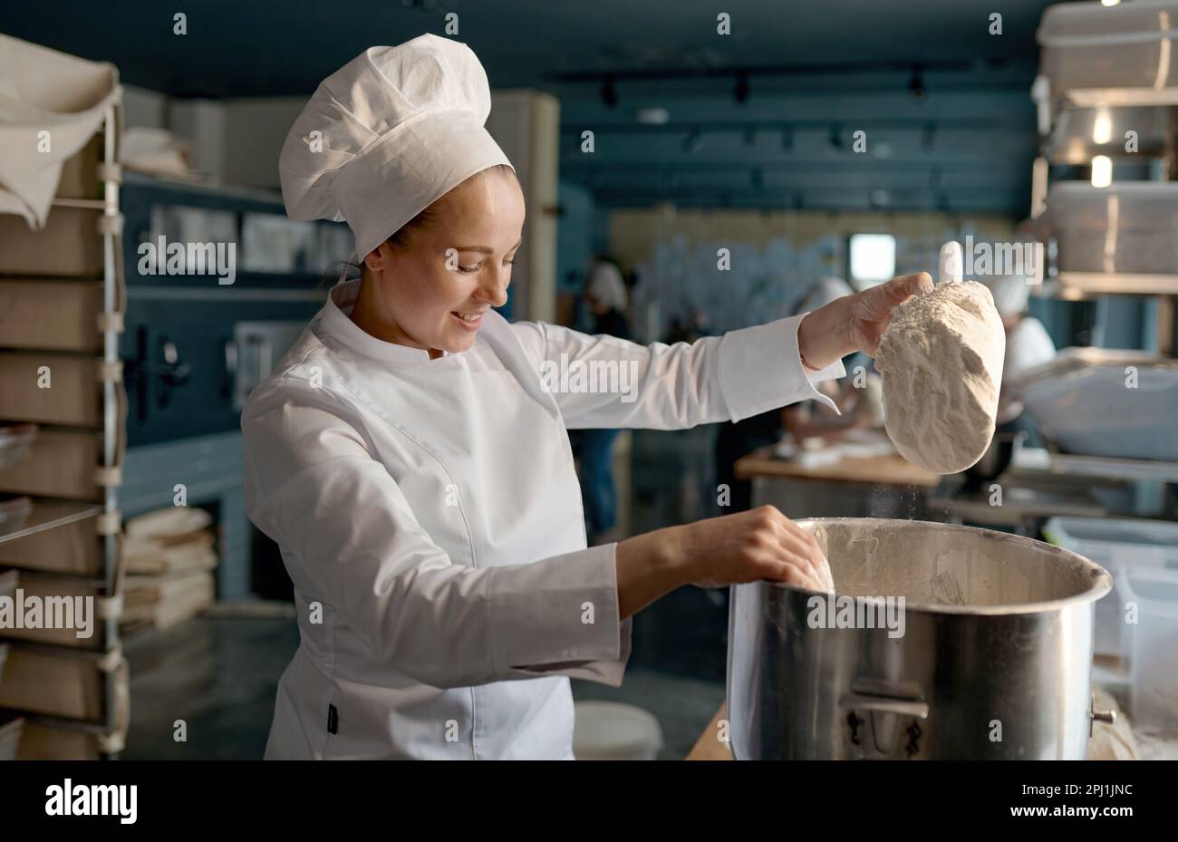 Female confectioner wearing white uniform putting flour into big metal ...