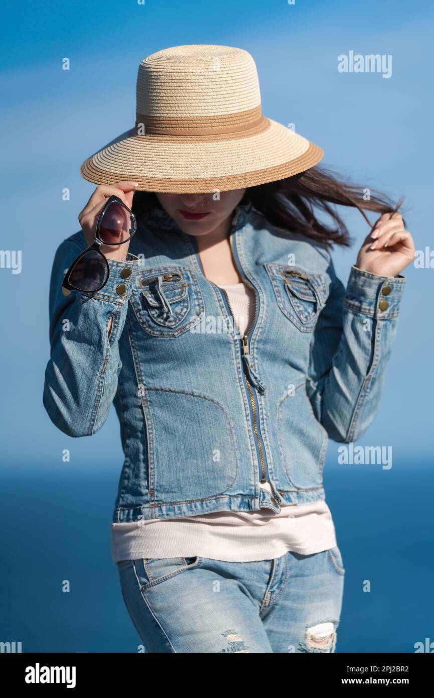 Portrait of female in denim jacket and jeans hiding with straw hat over ...