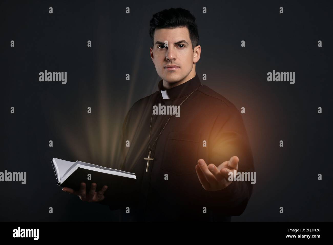 Priest in cassock holding Bible with holy light on black background ...