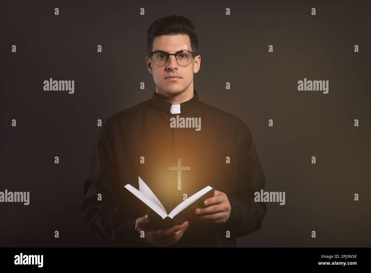 Priest in cassock holding Bible with holy light on black background ...