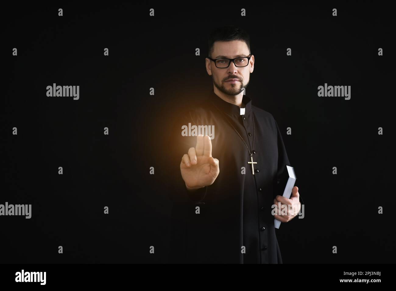 Priest with Bible making blessing gesture and holy light on dark ...
